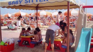 Cena de praia com área de serviço de 'BABY SITTER', incluindo mesas coloridas e um escorrega.