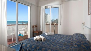 Quarto de hotel com uma cama, uma cadeira, uma mesinha e varanda com vista para a praia e o oceano.