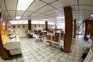 Uma ampla sala de jantar com pé direito alto, piso ladrilhado e diversas mesas e cadeiras.