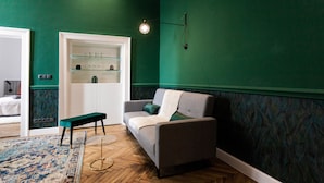 Quarto com parede verde, sofá cinza com almofada verde, piso de madeira em padrão herringbone e tapete estampado.