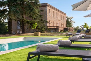 Área da piscina com espreguiçadeiras e toalhas, um edifício de pedra ao fundo e céu claro.