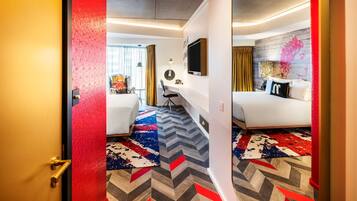 Quarto de hotel com cama, escrivaninha, cadeira, televisão e espelho.