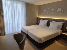 Quarto de hotel com duas camas, uma escrivaninha, uma cadeira e uma mesa de cabeceira com abajur.