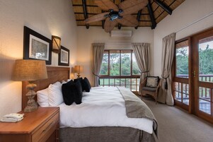 Quarto com cama grande, duas luminárias de cabeceira, mesa de cabeceira de madeira, uma cadeira junto à janela e um ventilador de teto.