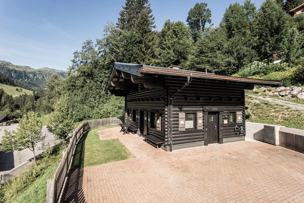 Uma cabana de toras com um pátio de tijolos, cercada por uma cerca de madeira, com montanhas e árvores ao fundo.