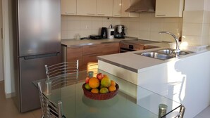 Uma cozinha moderna com geladeira de aço inoxidável, armários de madeira, mesa de jantar de vidro e uma cesta de frutas, além de forno embutido.