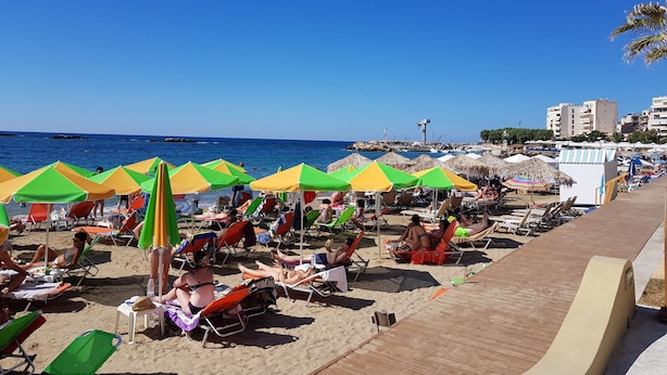 Uma praia com vários guarda-sóis coloridos e espreguiçadeiras, um calçadão de madeira e um céu azul límpido.