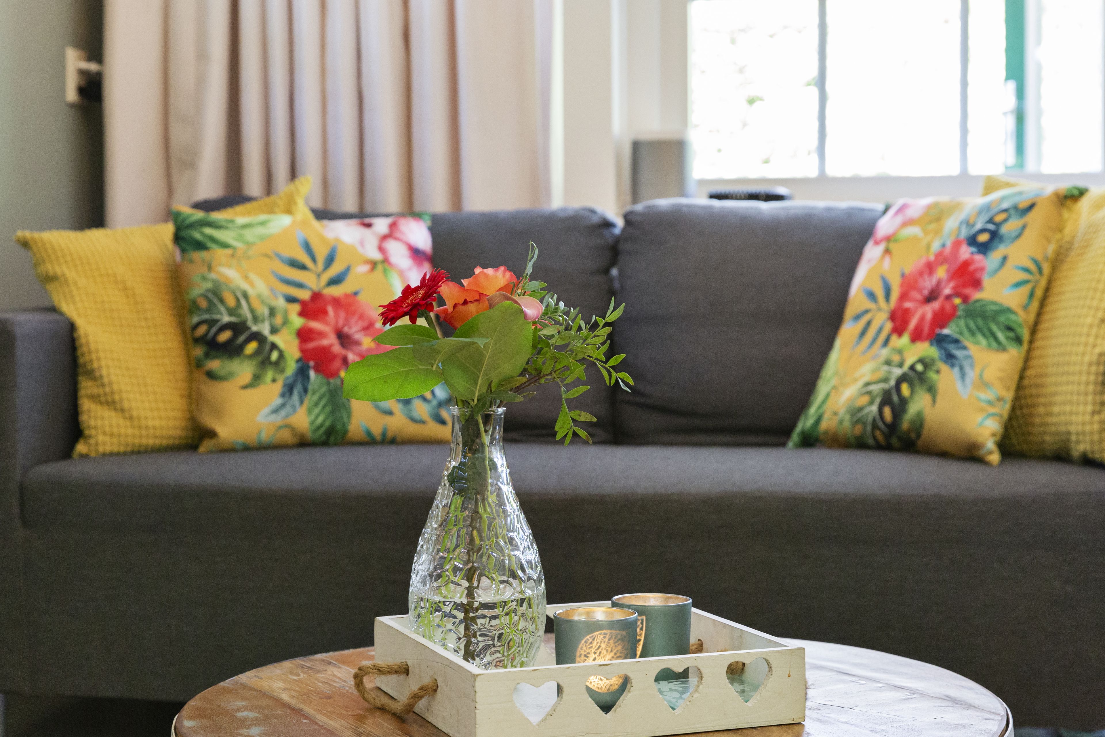 Sala de estar com sofá cinza, almofadas florais, um vaso com flores e um tabuleiro com velas.