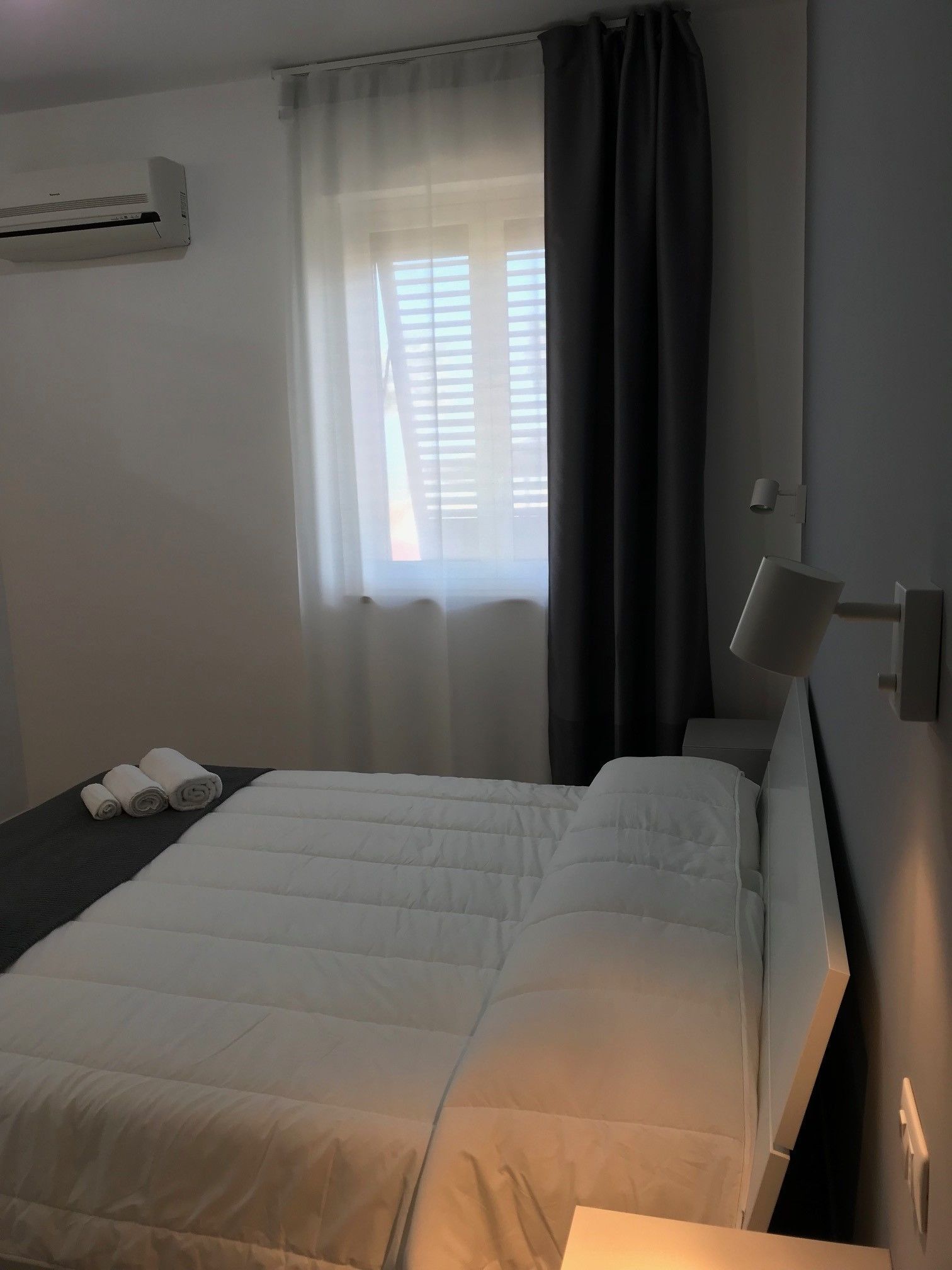 Cama bem arrumada com edredom branco e travesseiros, mesa de cabeceira com abajur, janela com persianas e ar condicionado na parede.