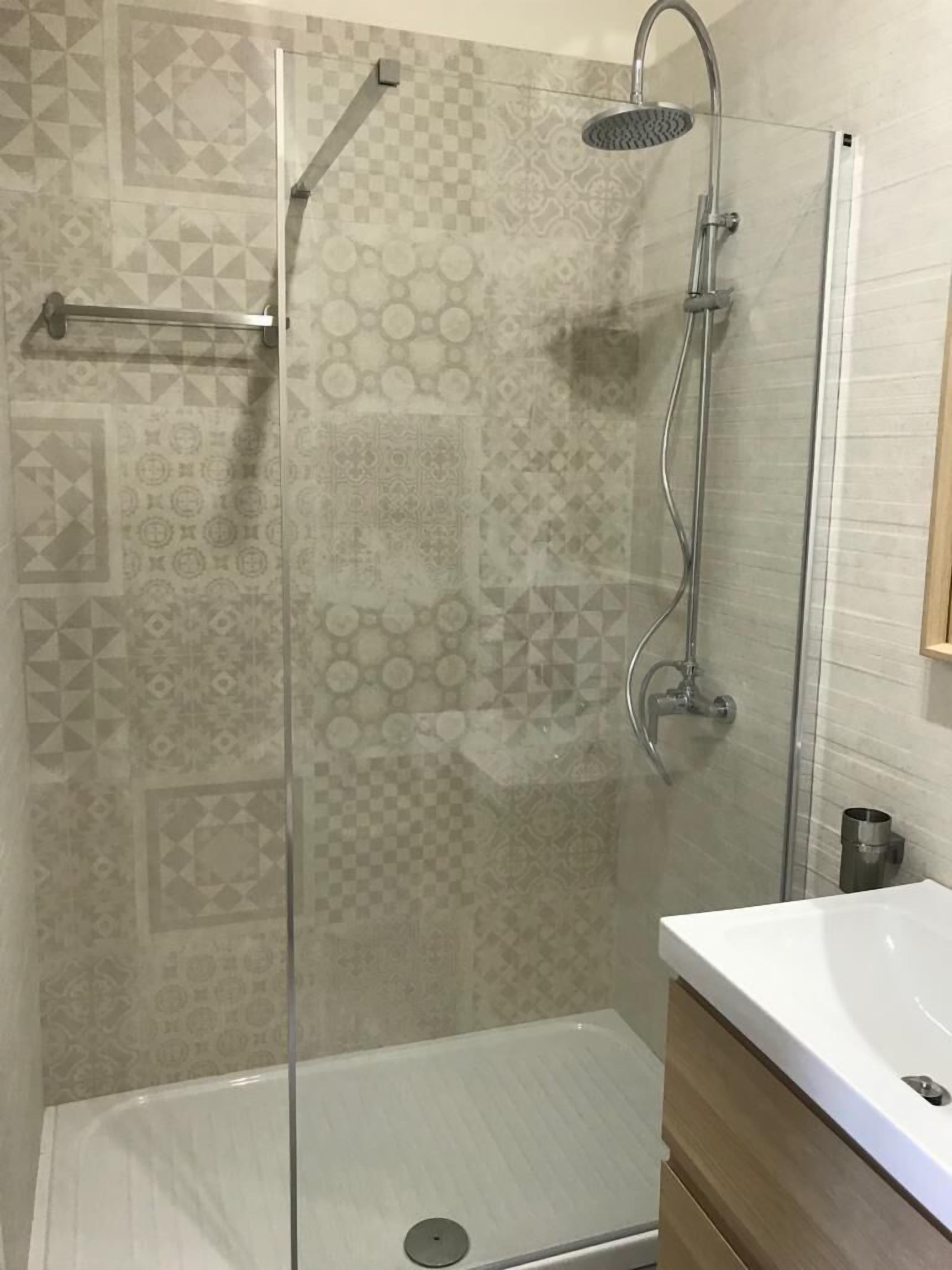 Um chuveiro com porta de vidro, uma pia branca e azulejos estampados nas paredes.