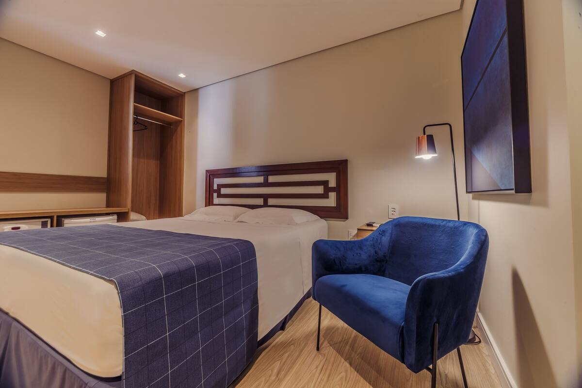 Quarto de hotel com cama, uma cadeira, guarda-roupa e mesa de cabeceira.