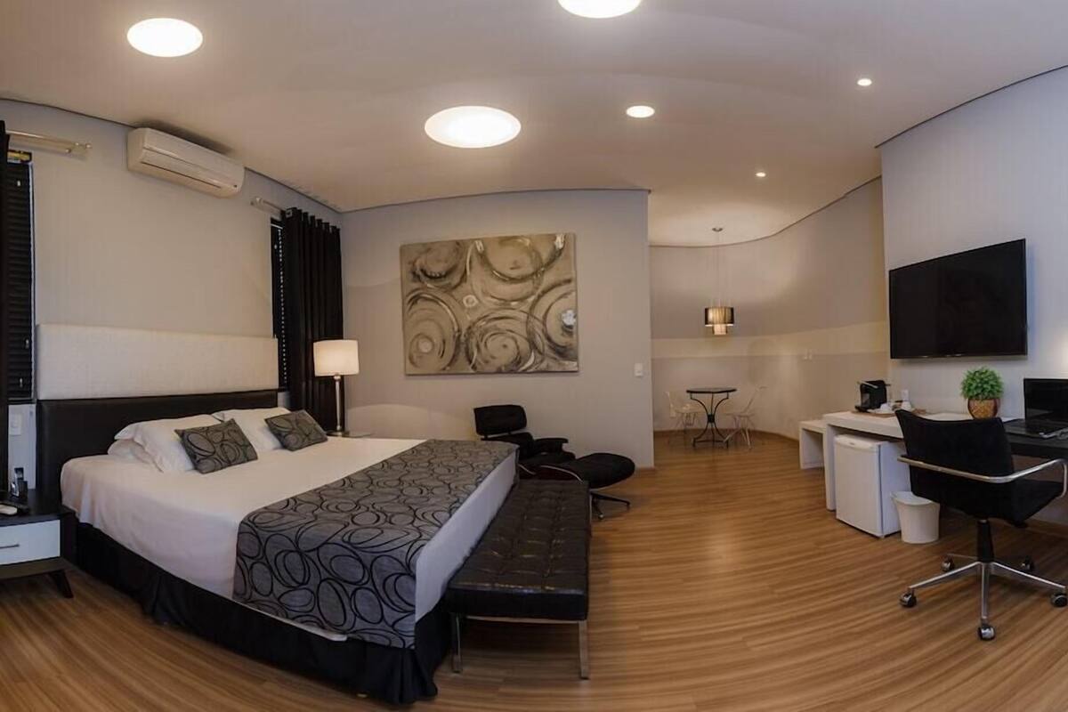 Quarto de hotel moderno com uma cama grande, uma escrivaninha, uma cadeira, uma televisão e um quadro na parede.