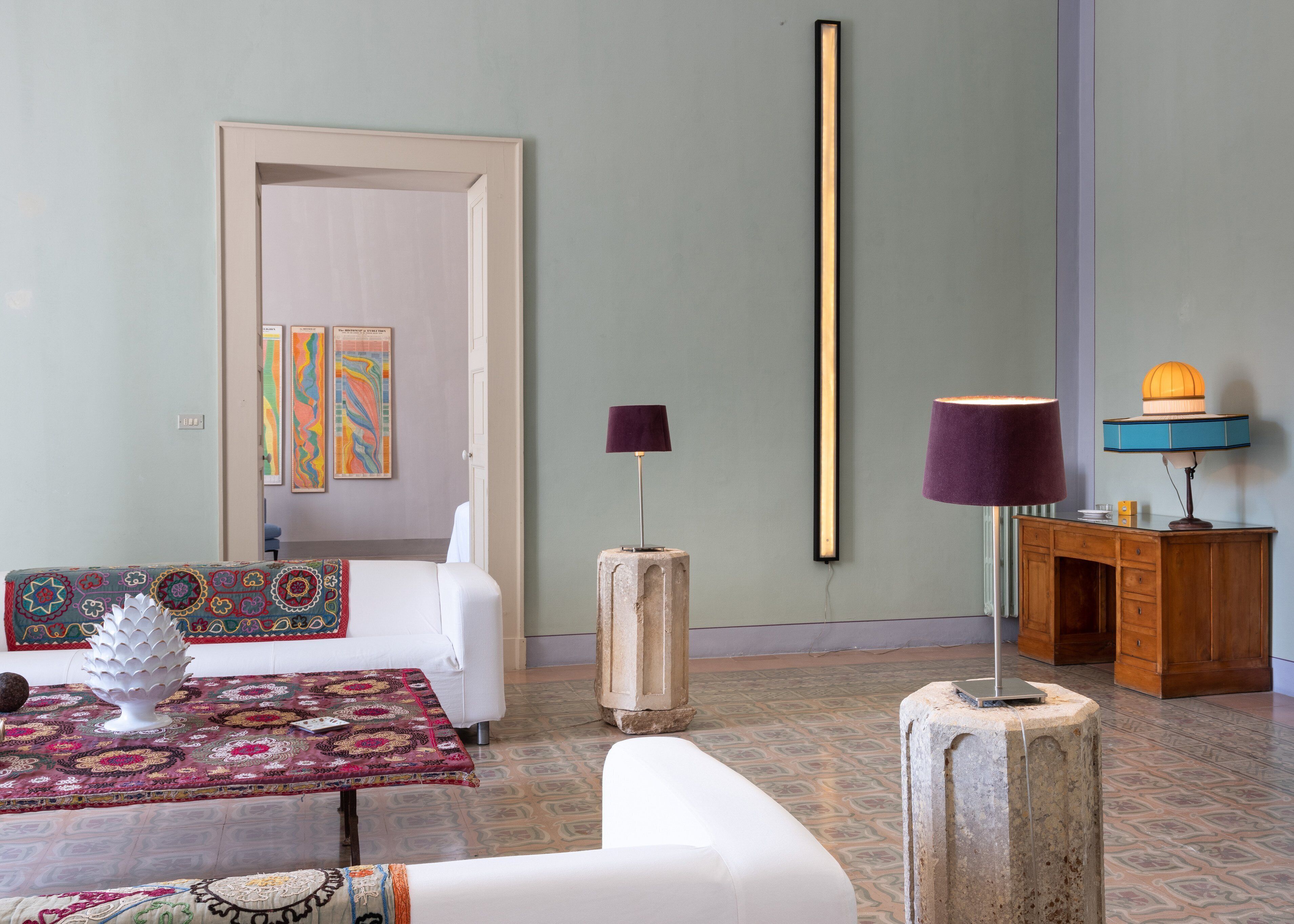Uma sala de estar moderna com um sofá, uma mesa de centro, uma luminária e um armário. Há uma porta que dá para outro quarto com obras de arte coloridas na parede.