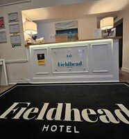 Bancada de recepção no Fieldhead Hotel, com carpete preto e texto branco, certificados emoldurados na parede e um quadro sobre a mesa.