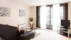 Uma sala de estar moderna com um sofá de couro preto, uma mesa de centro de vidro, uma televisão de tela plana em um móvel e janelas amplas com cortinas leves.