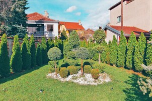 Um jardim bem cuidado, com sebes aparadas e uma árvore central, rodeado por um muro de tijolos e casas com telhados vermelhos.