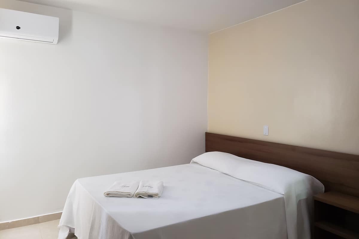 Uma cama pequena e bem arrumada, com colchão branco e cabeceira de madeira. Há um ar condicionado branco na parede, acima da cama.