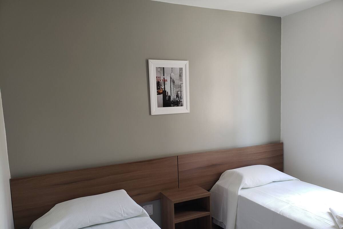 Quarto de hotel com duas camas de solteiro, uma pequena mesa de cabeceira de madeira e um quadro na parede.