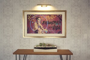 Obra de arte emoldurada com um design abstrato vibrante e texto, colocada acima de uma pequena mesa com uma planta.