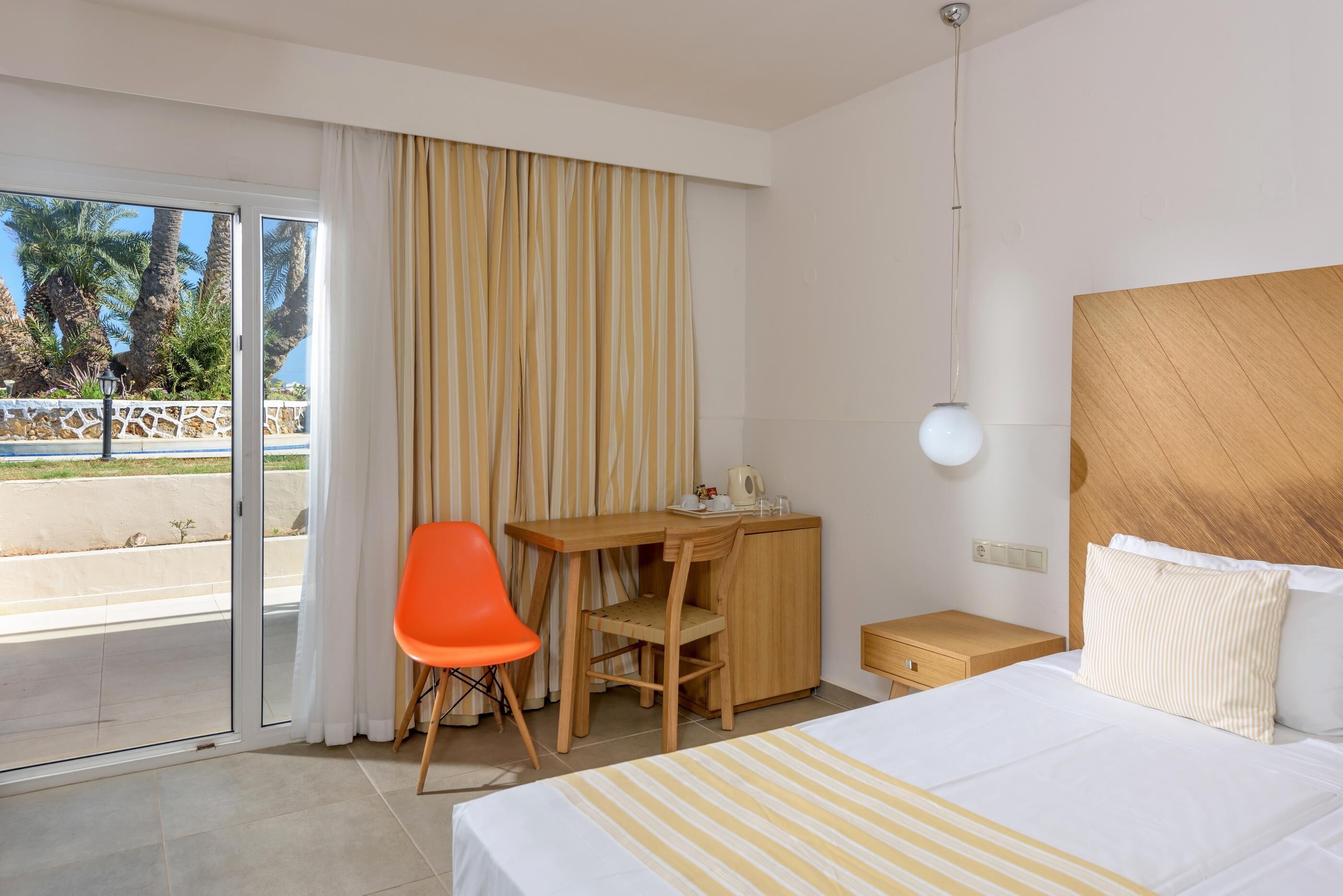 Quarto de hotel com uma cama, uma escrivaninha, uma cadeira laranja, uma cabeceira de madeira e vista para palmeiras através de uma porta deslizante.
