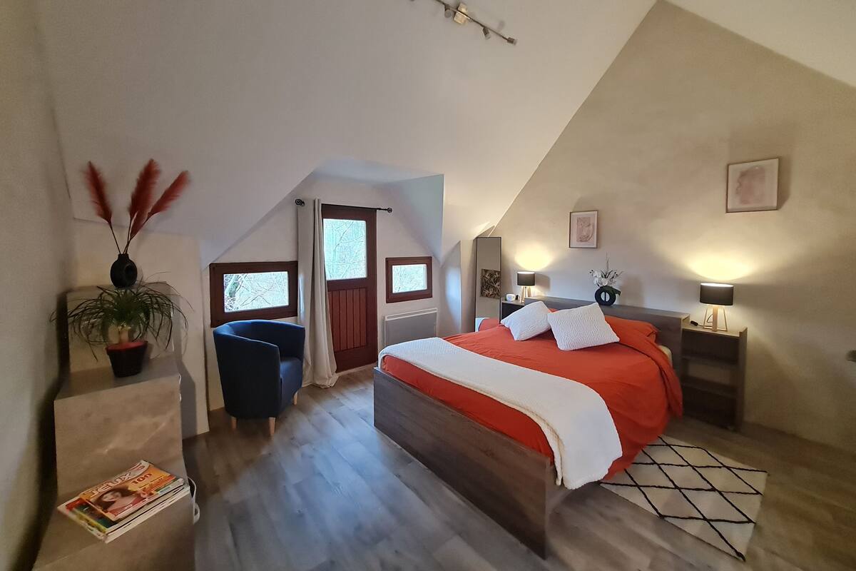 Um quarto com teto inclinado, uma cama com roupa de cama branca e laranja, uma cadeira azul, uma mesa de cabeceira com abajur e uma janela com vista para árvores.