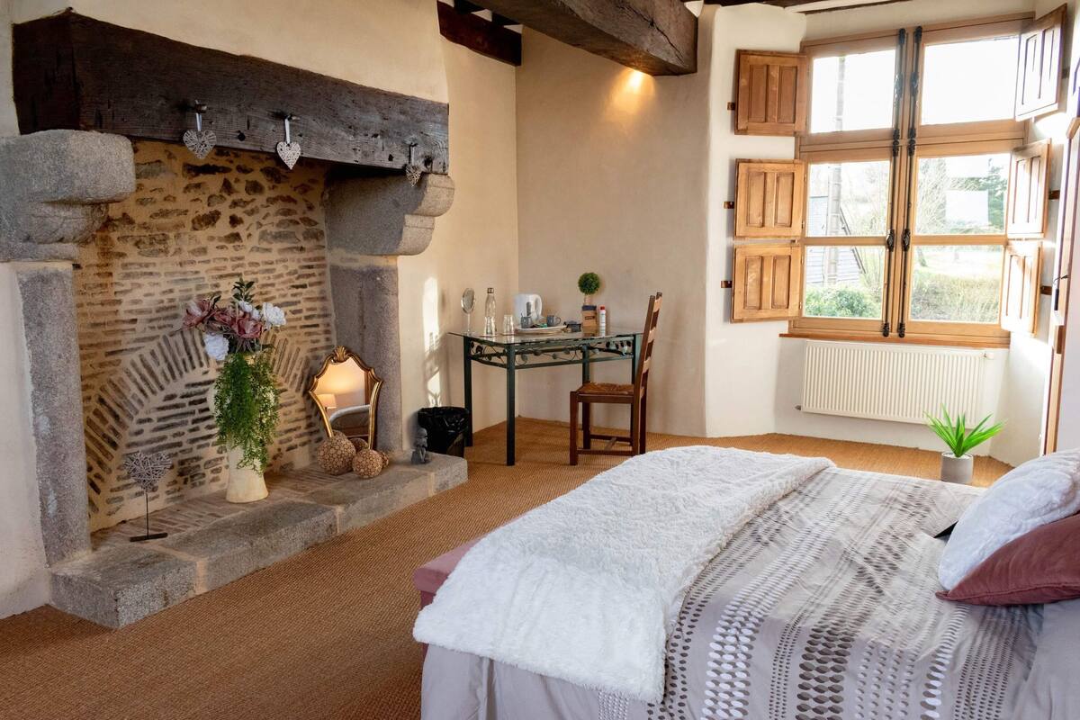 Um quarto acolhedor com lareira de pedra, uma cama com cobertor branco, uma mesa pequena com uma cadeira e uma janela com persianas de madeira.