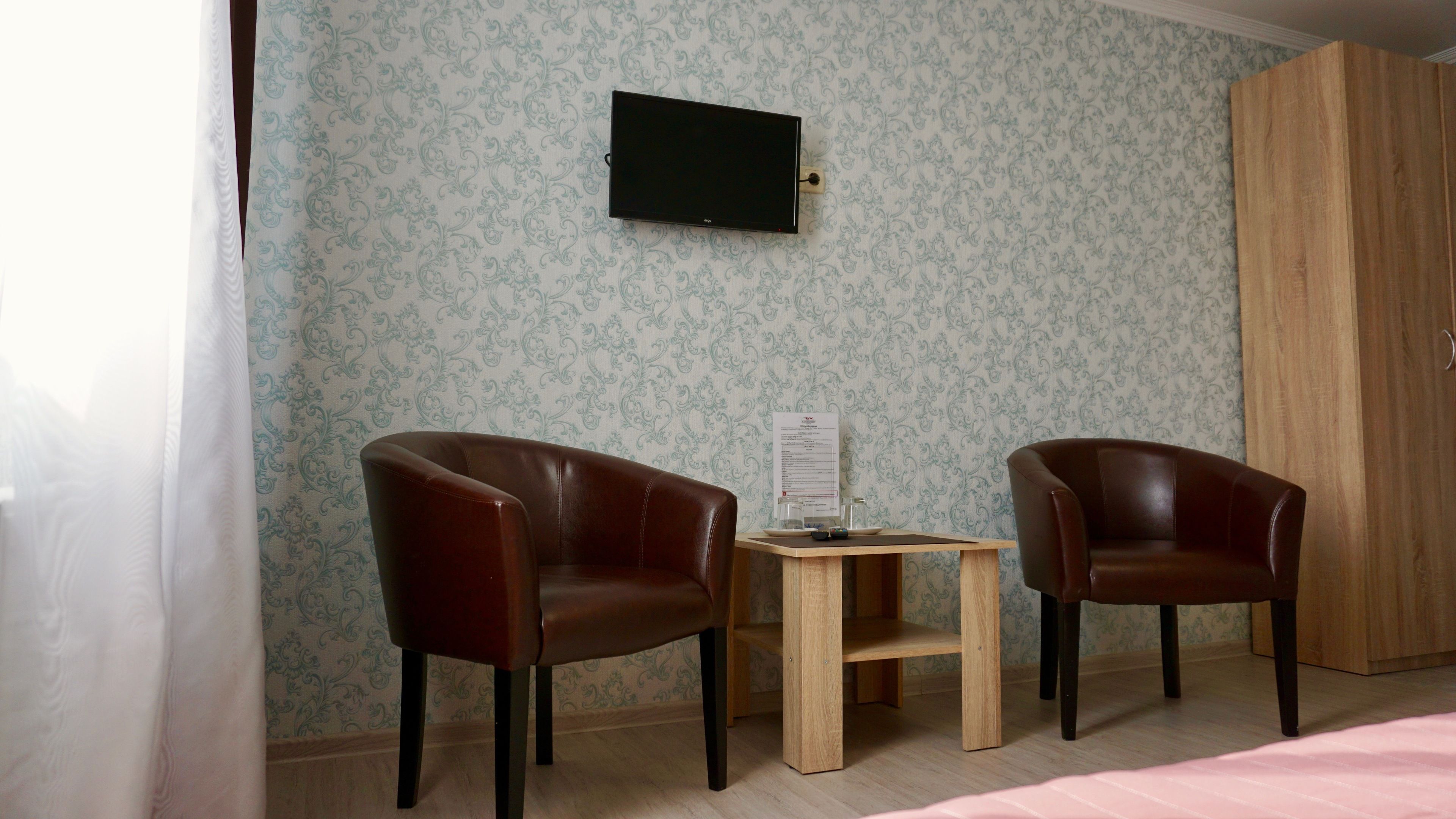 Quarto com duas poltronas de couro, uma mesa de madeira pequena, televisão de tela plana na parede e um guarda-roupa.