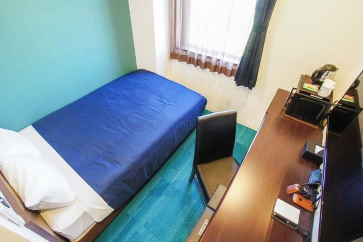 Um quarto pequeno, com cama de solteiro, colcha azul, escrivaninha de madeira, uma cadeira e uma mesinha com telefone e uma xícara.