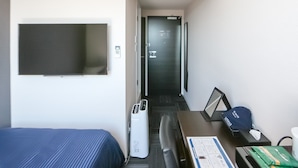 Quarto de hotel com uma cama, uma escrivaninha com computador, uma televisão na parede e uma porta para o corredor.