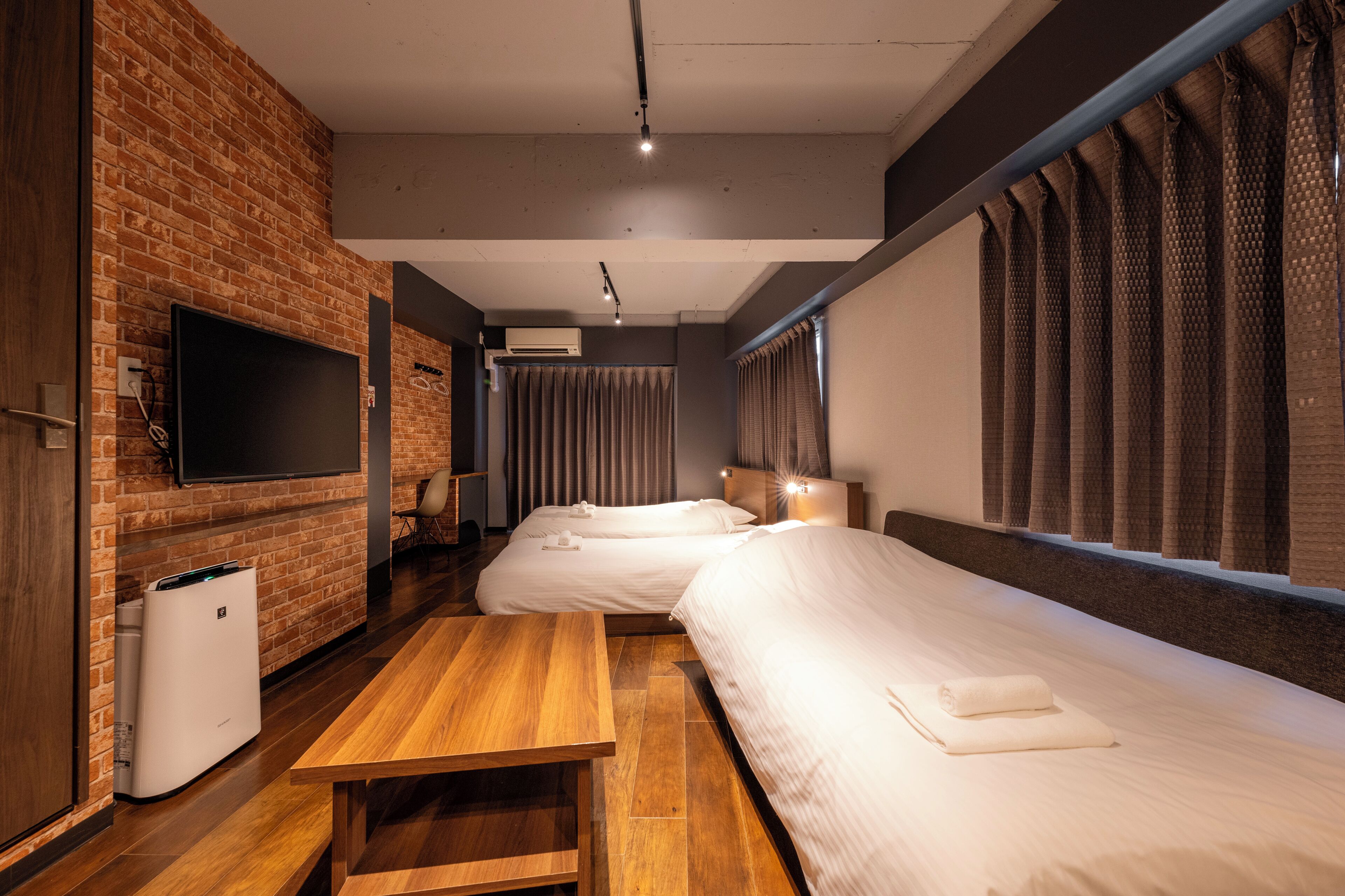 Quarto de hotel com parede de tijolos, televisão de tela plana, mesa de madeira, duas camas e janela com cortinas.