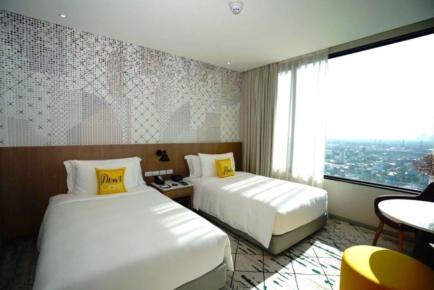 Quarto de hotel com duas camas, uma escrivaninha, uma cadeira e uma janela grande com vista para a cidade.