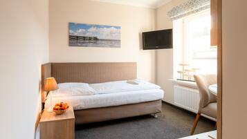 Quarto de hotel com cama, mesa de cabeceira com abajur e tigela de frutas, televisão e janela com vista para o mar.
