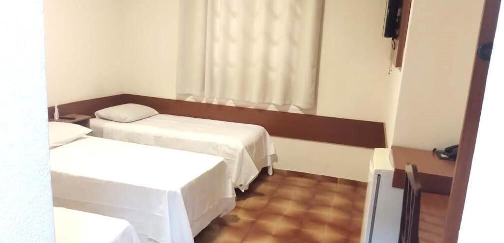 Quarto com duas camas de solteiro, piso de madeira, janela com cortinas e uma pequena mesa com uma cadeira.