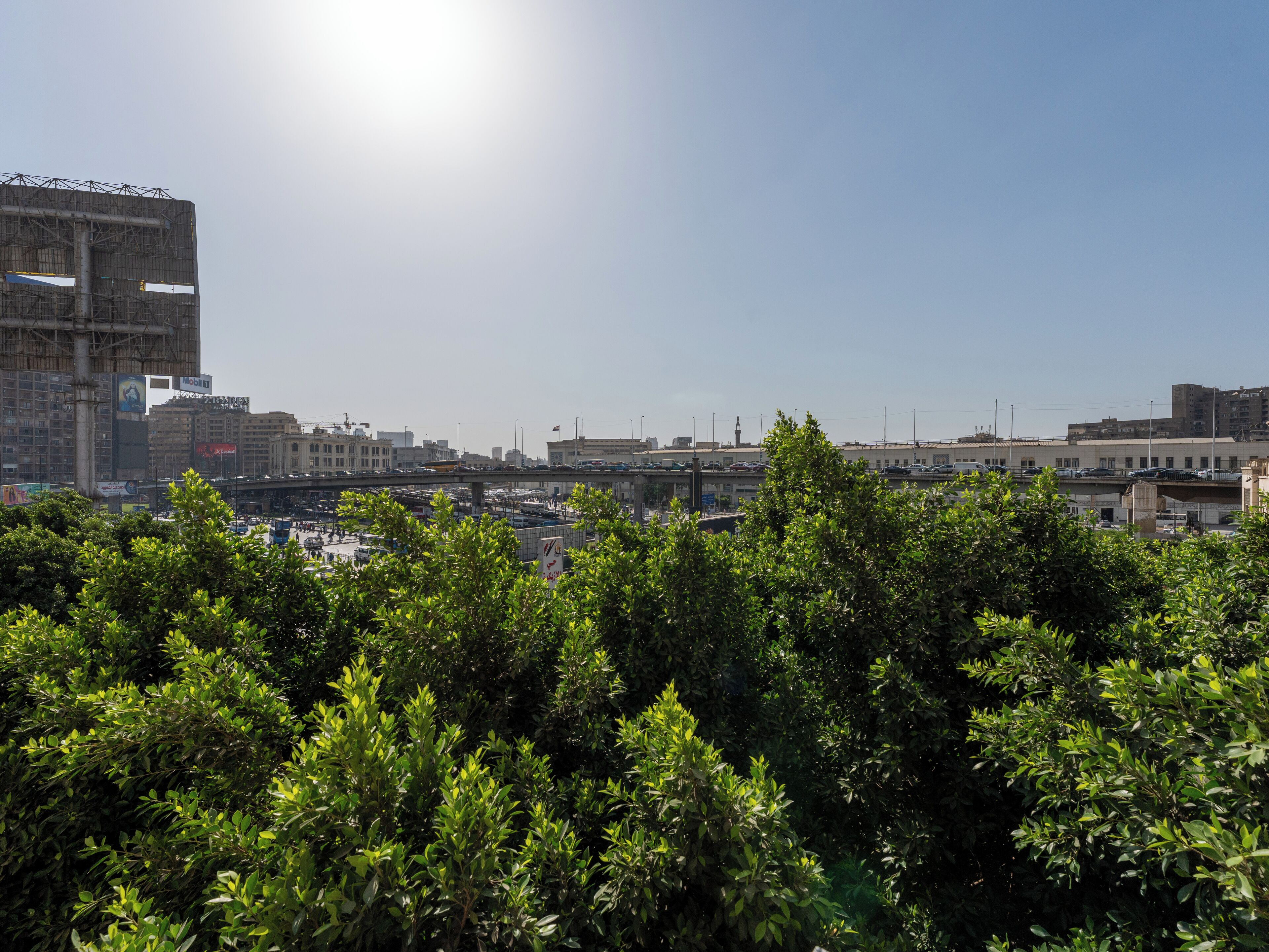 Paisagem urbana com um edifício em construção em destaque, rua movimentada com veículos e árvores verdes exuberantes em primeiro plano.