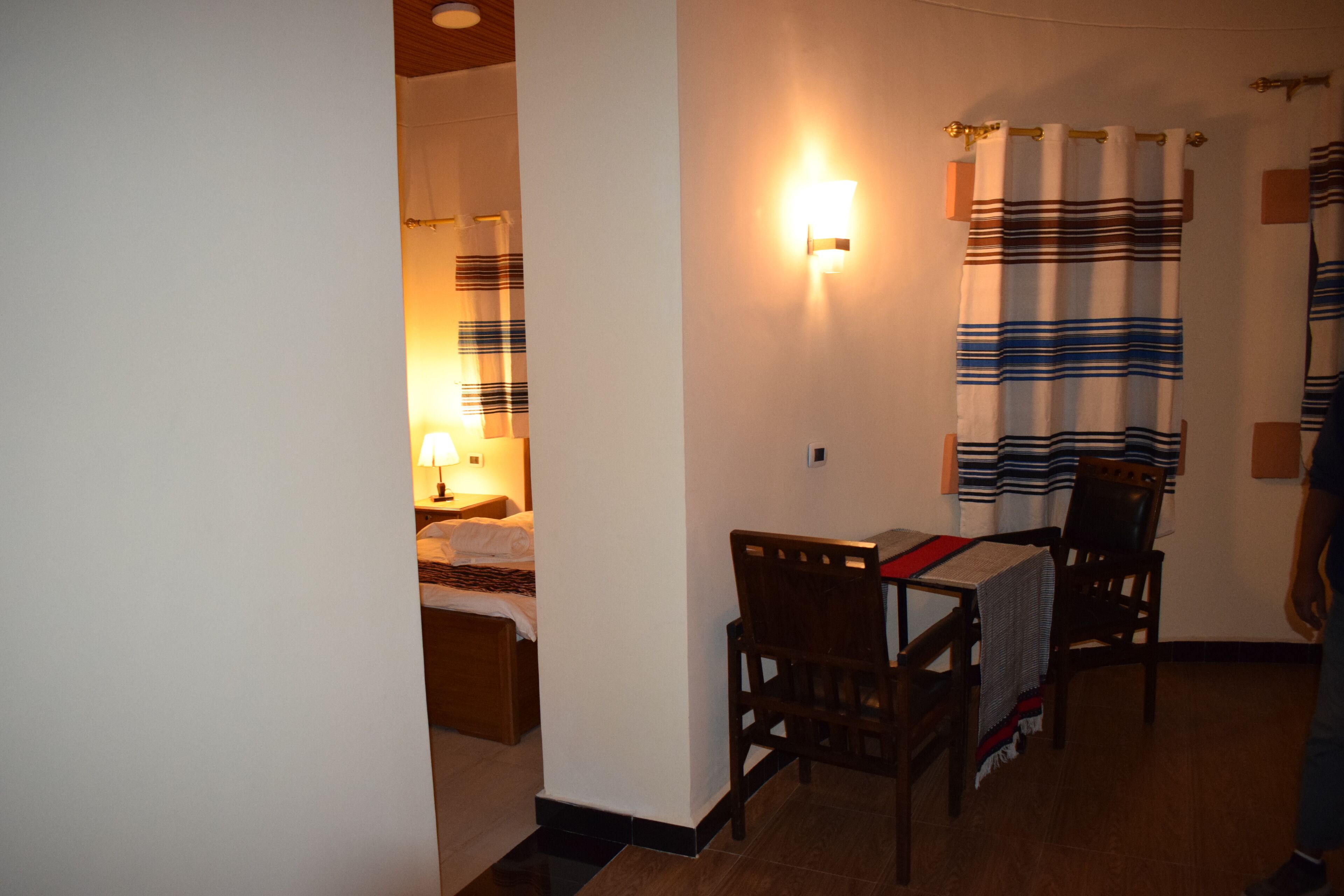 Quarto de hotel com cama, mesa de cabeceira, mesa com cadeiras e janela com cortinas listradas.