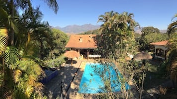 Uma casa com piscina, rodeada por vegetação tropical e montanhas ao fundo.