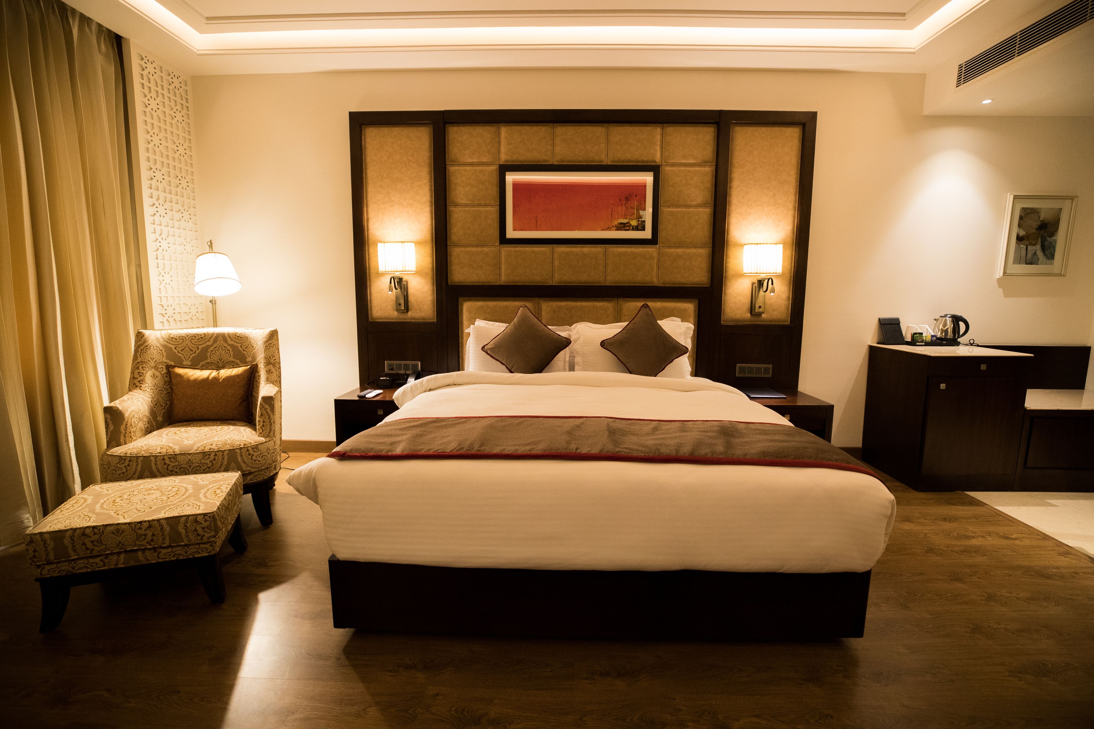 Quarto de hotel com uma cama grande, duas luminárias de cabeceira, uma cadeira e uma escrivaninha com chaleira.