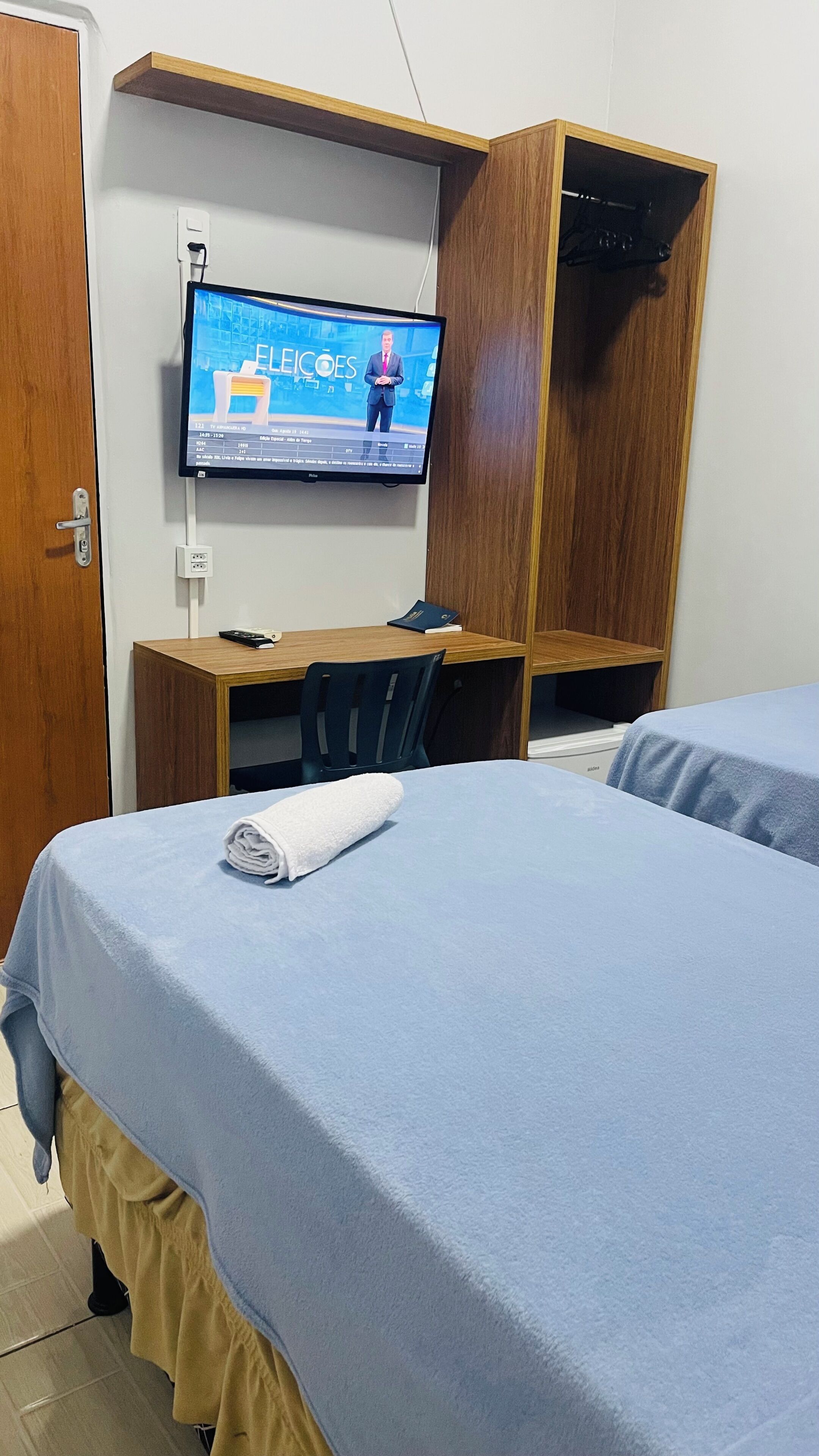 Quarto de hotel com cama, escrivaninha com computador, televisão na parede e guarda-roupa.