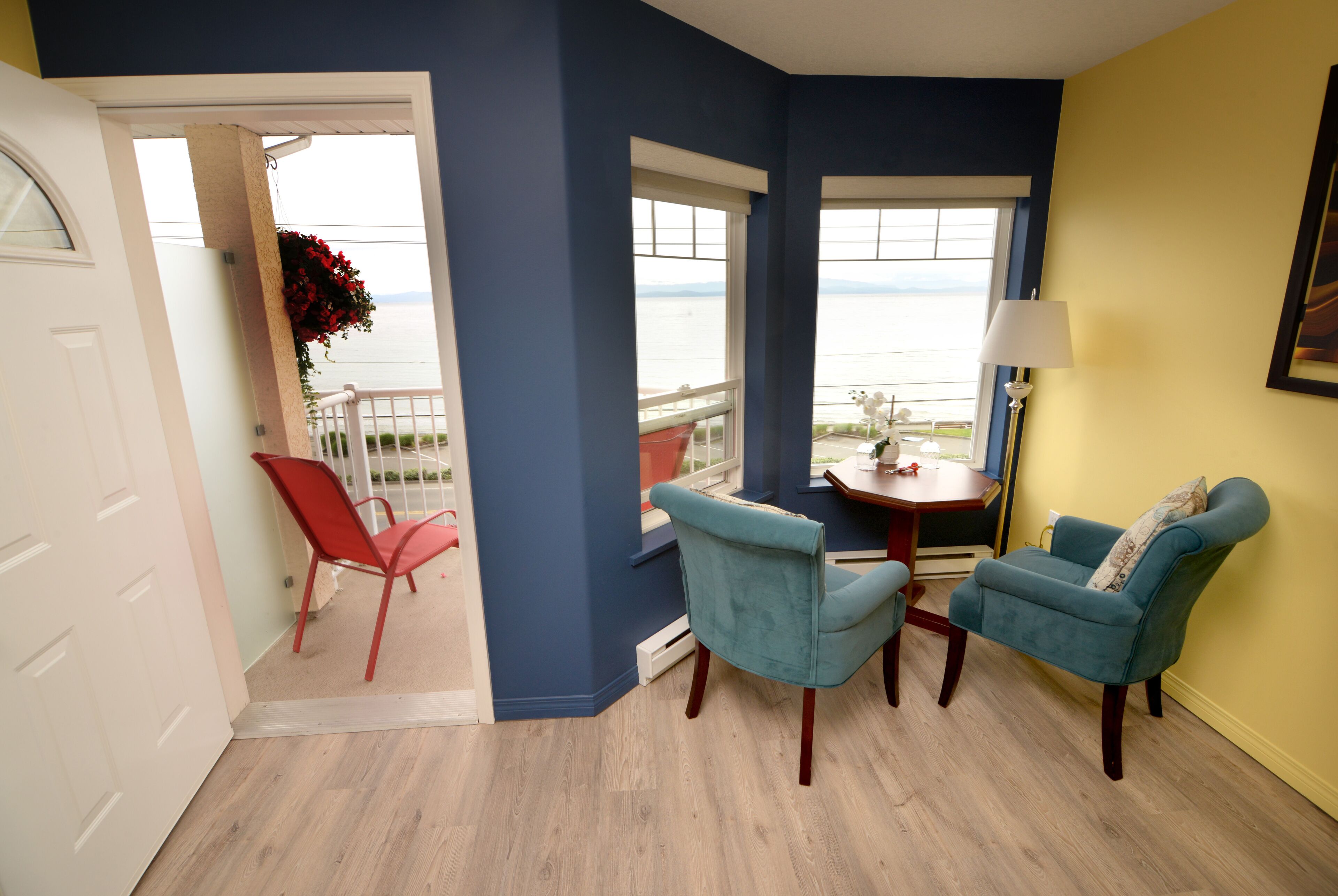 Quarto com vista para o mar, com duas cadeiras, uma mesa e um abajur.