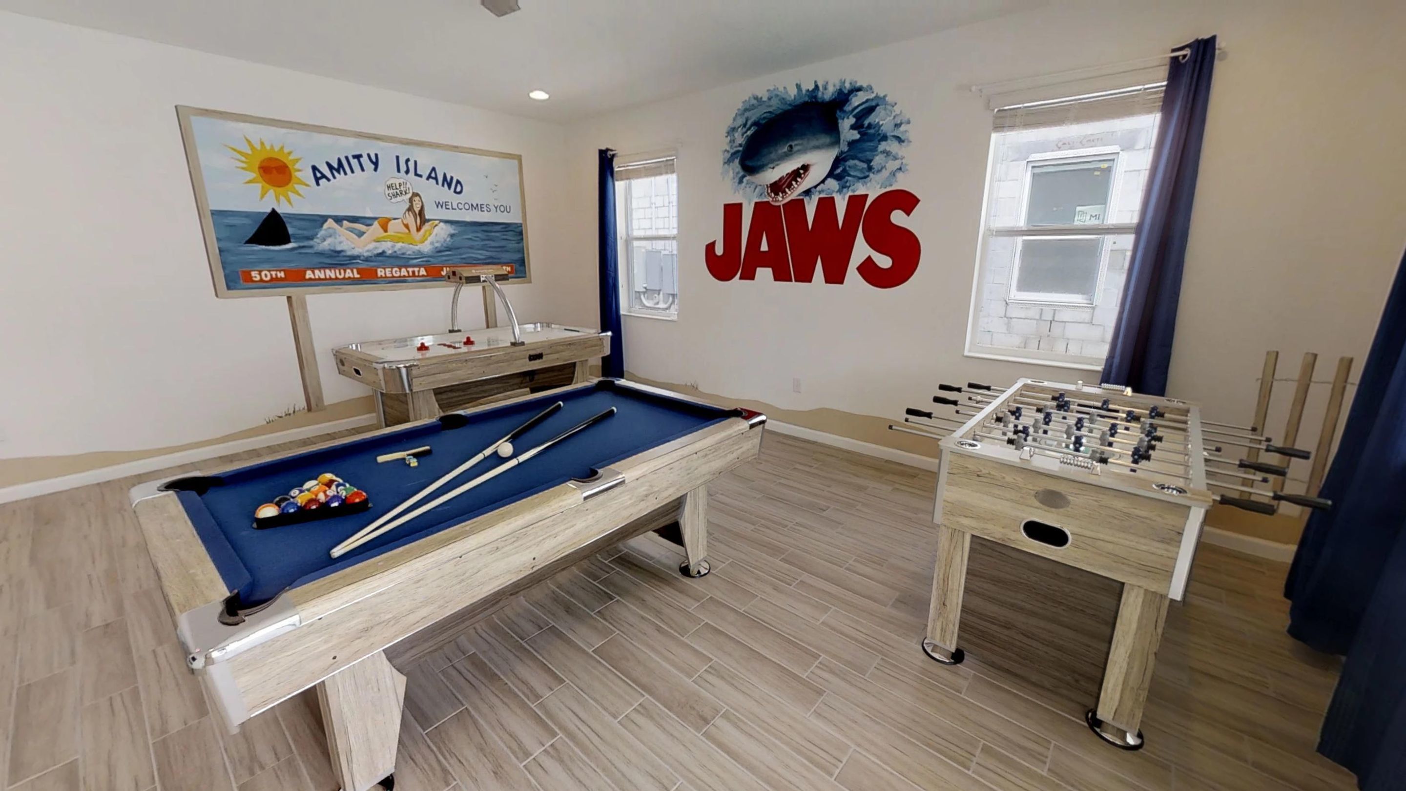 Quarto com mesa de bilhar, mesa de futebol e parede com ilustração de tubarão e texto.