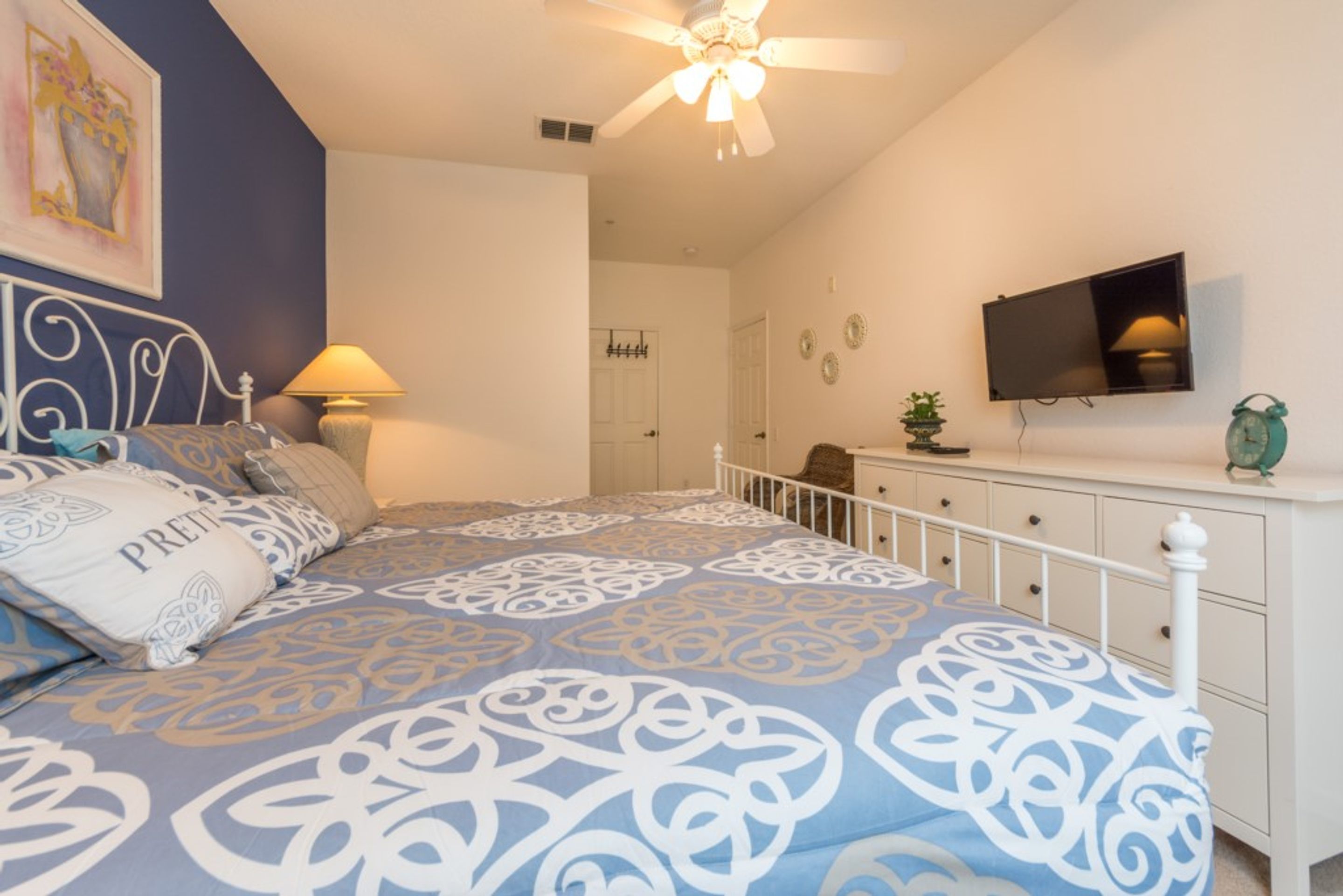 Quarto com cama, ventilador de teto, televisão, cômoda e uma porta ao fundo.