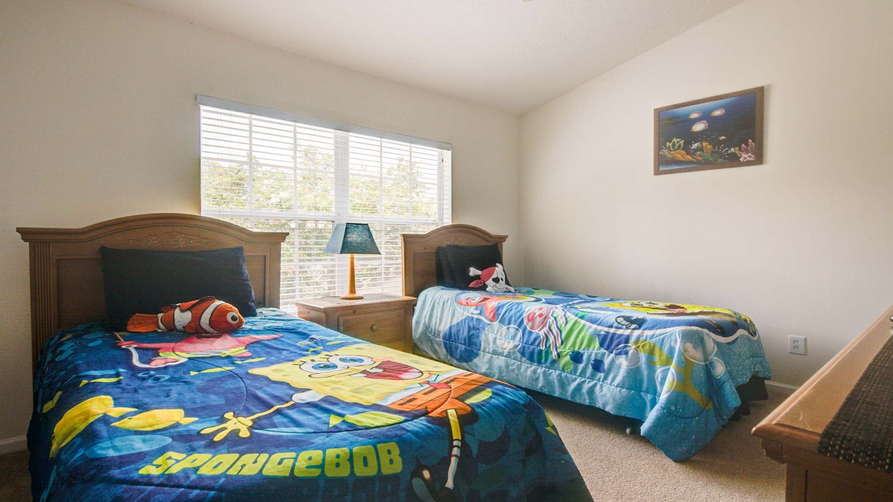 Quarto com duas camas, cada uma com colcha azul com tema do Bob Esponja. Há uma janela com persianas, uma mesa de cabeceira com abajur e um quadro fixado na parede.