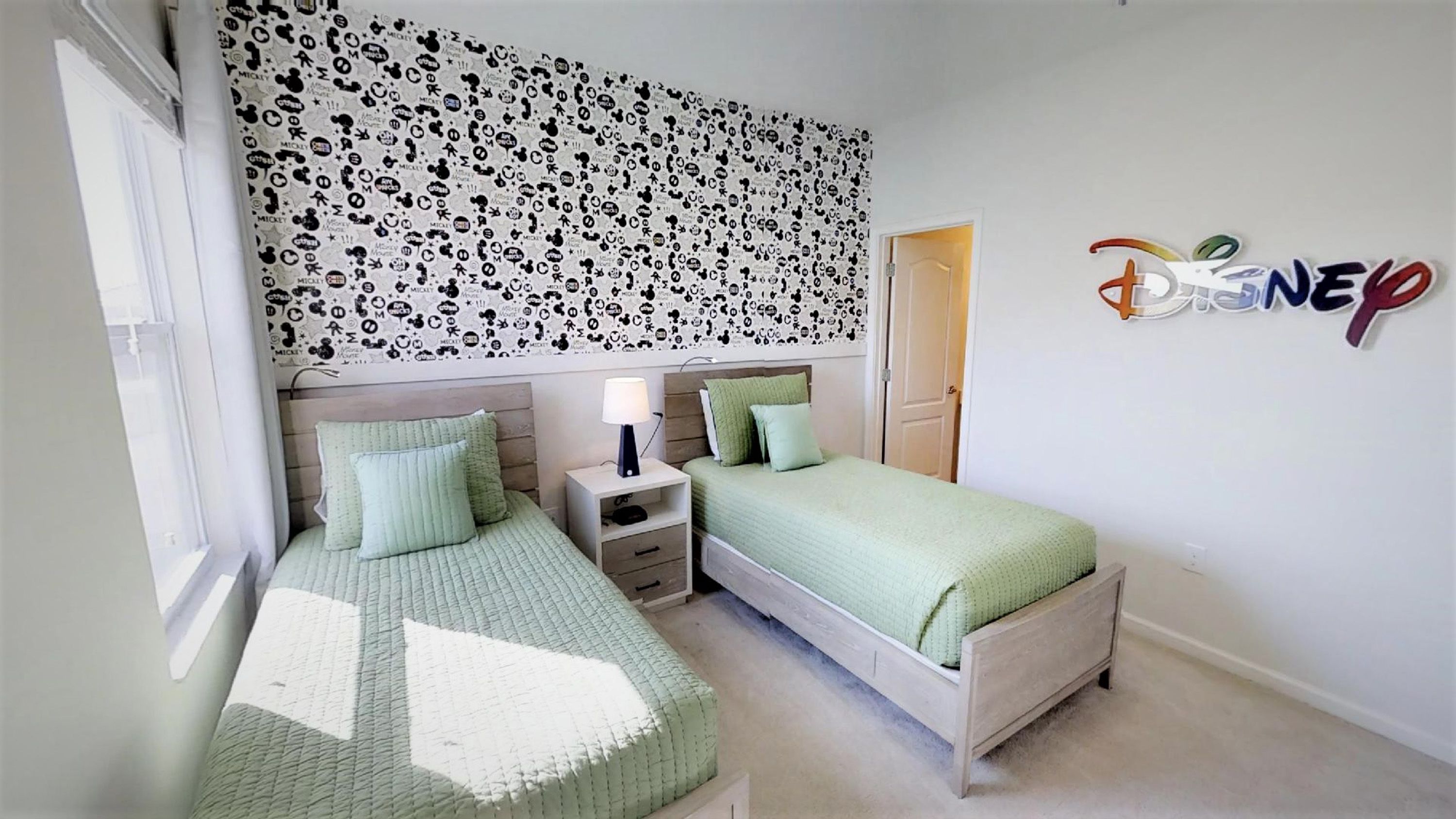 Quarto com duas camas, uma mesa de cabeceira, uma luminária e parede com padrão do Mickey Mouse.