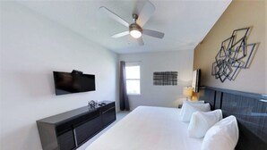 Quarto de hotel moderno com uma cama grande, ventilador de teto, TV de tela plana na parede, mesa de console escura e uma janela com cortinas.