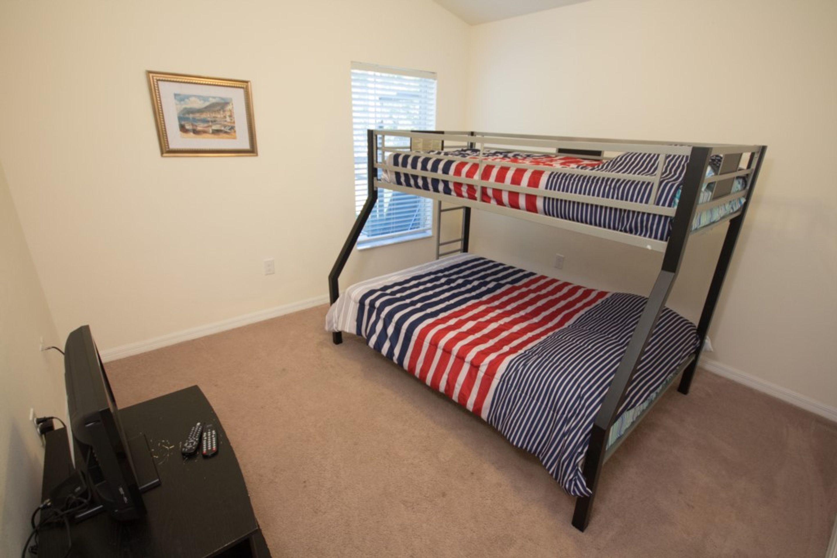 Quarto com beliche, um quadro na parede, escrivaninha com computador e controles remotos, e janela com persianas.