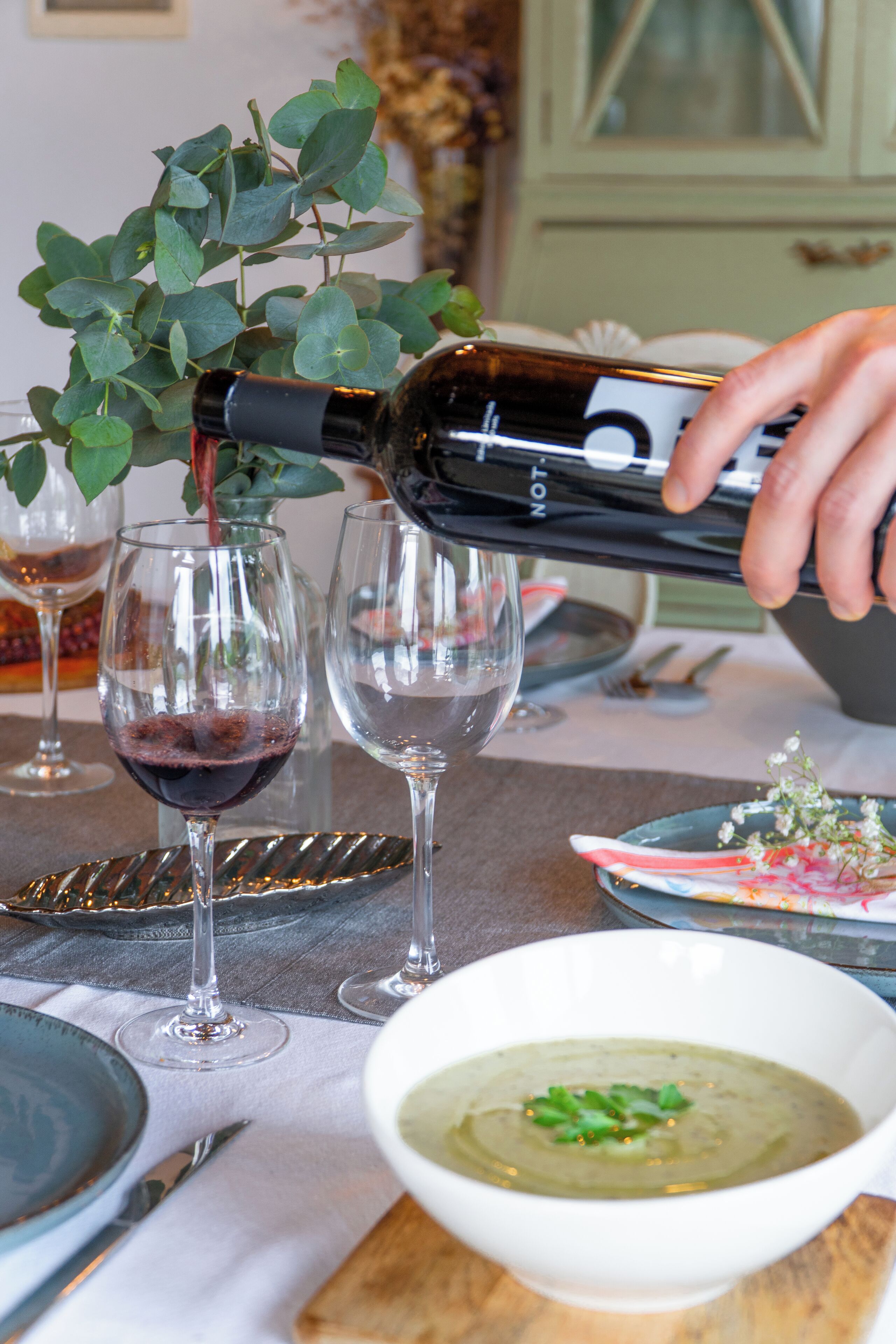 Uma mesa de jantar com copos de vinho, uma tigela de sopa e uma garrafa sendo servida em um dos copos.