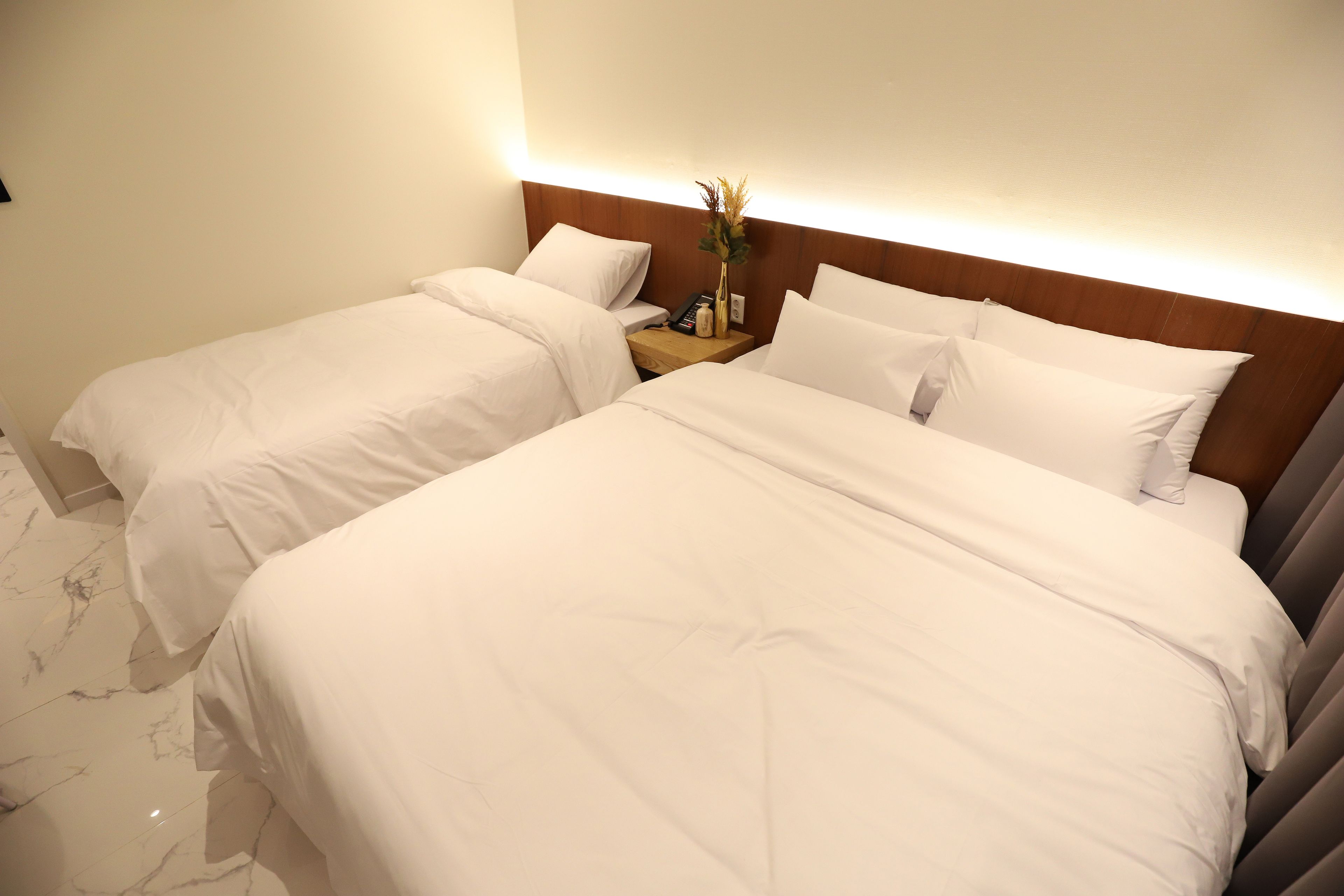 Quarto de hotel com duas camas de solteiro, cabeceira de madeira, mesa de cabeceira com um vaso e piso de mármore.