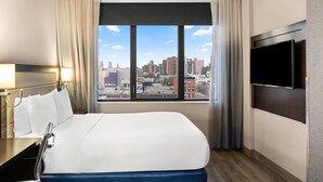 Quarto de hotel com uma cama grande, televisão fixada na parede e janela com vista para a cidade.