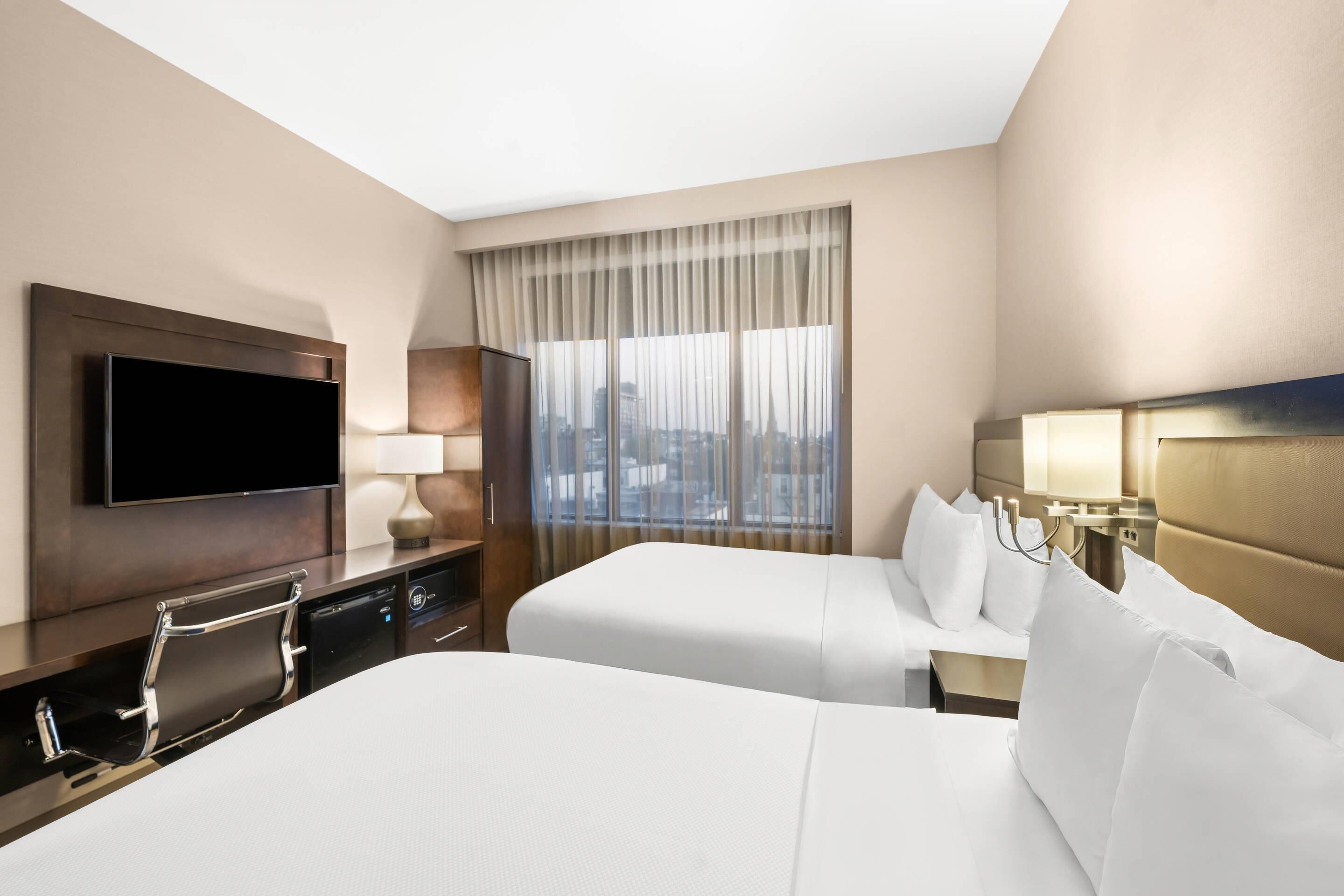 Quarto de hotel com uma cama grande, televisão presa na parede, escrivaninha com luminária e janela com cortinas.