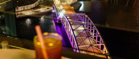 Vista de uma ponte iluminada em roxo, uma mesa de vidro com uma bebida e um horizonte urbano ao fundo.